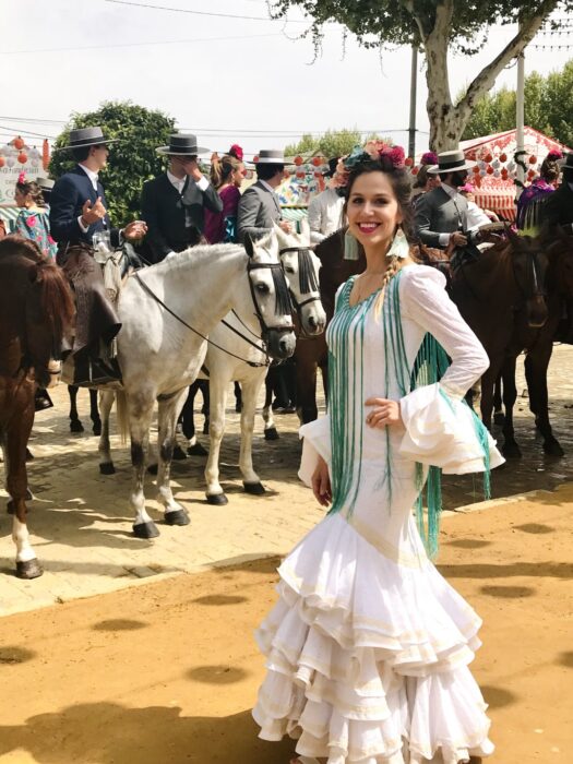 Emily dressed in a white dress posing in front of horses and riders at a fiesta in Seville