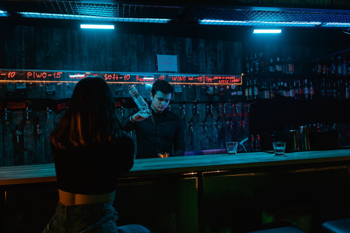 Bartender pouring a drink under neon bar lights with patron seated at counter