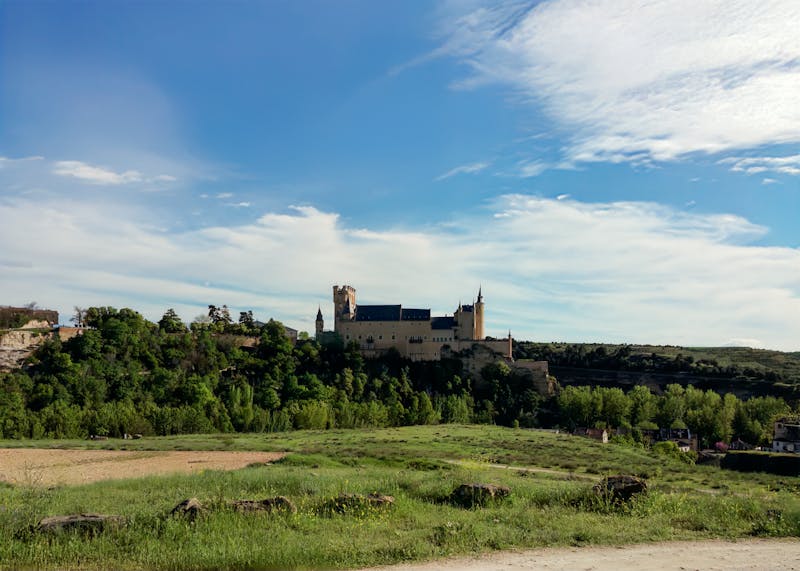 The Alcazar of Segovia sitting on top of a rocky hill surrounded by green vegetation and blue sky
