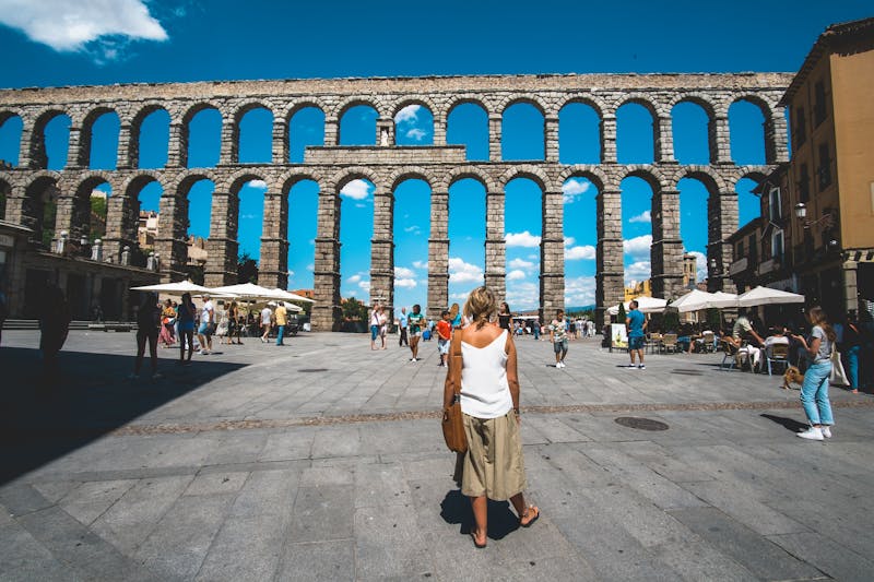 Tourists gathered at the base of the Roman Aqueduct in Segovia, with cafes and buildings lining the plaza