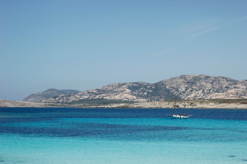 A sailing yacht on turquoise Mediterranean water near a rocky Sardinian coastline