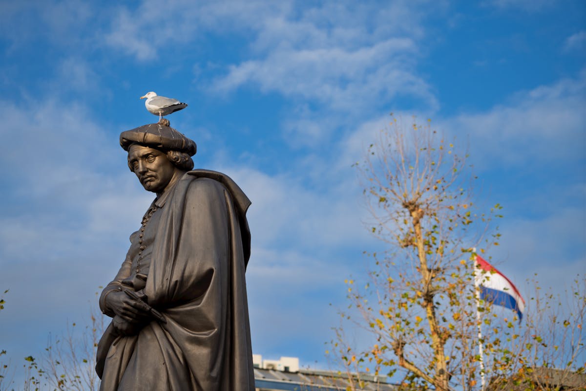 A seagull sits on the head of the Rembrandt statue with a Dutch flag waving in the background