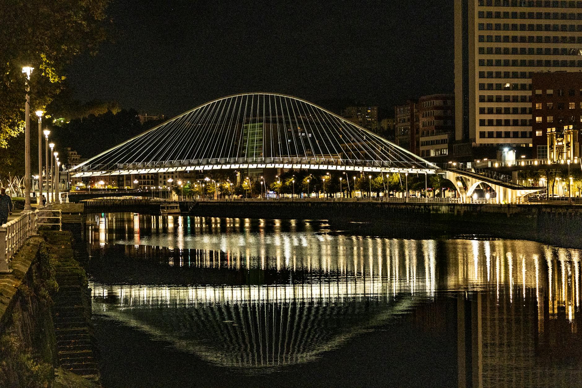 The illuminated Zubizuri pedestrian bridge in Bilbao Spain at night reflecting on the Nervion River