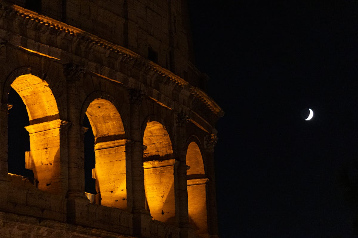 The Colosseum illuminated at night with a crescent moon above Rome