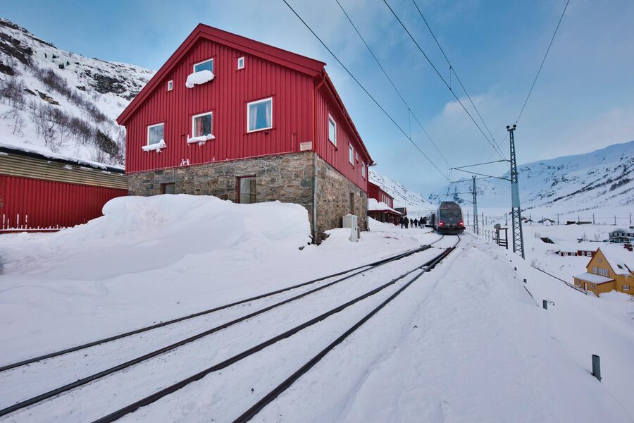 A red house beside train tracks in a snowy winter landscape with mountains in the background.