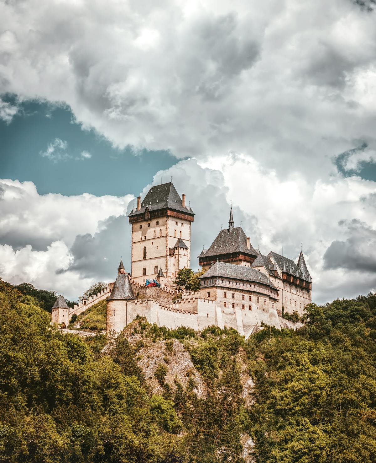 Medieval castle perched on a hilltop surrounded by lush greenery under dramatic clouds