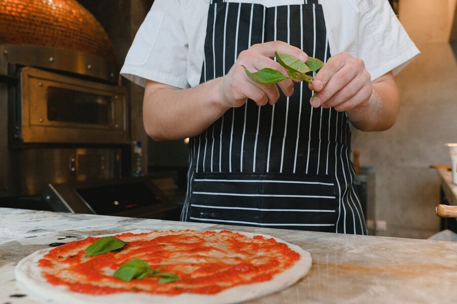 Chef in apron adding fresh basil to tomato sauce pizza in kitchen