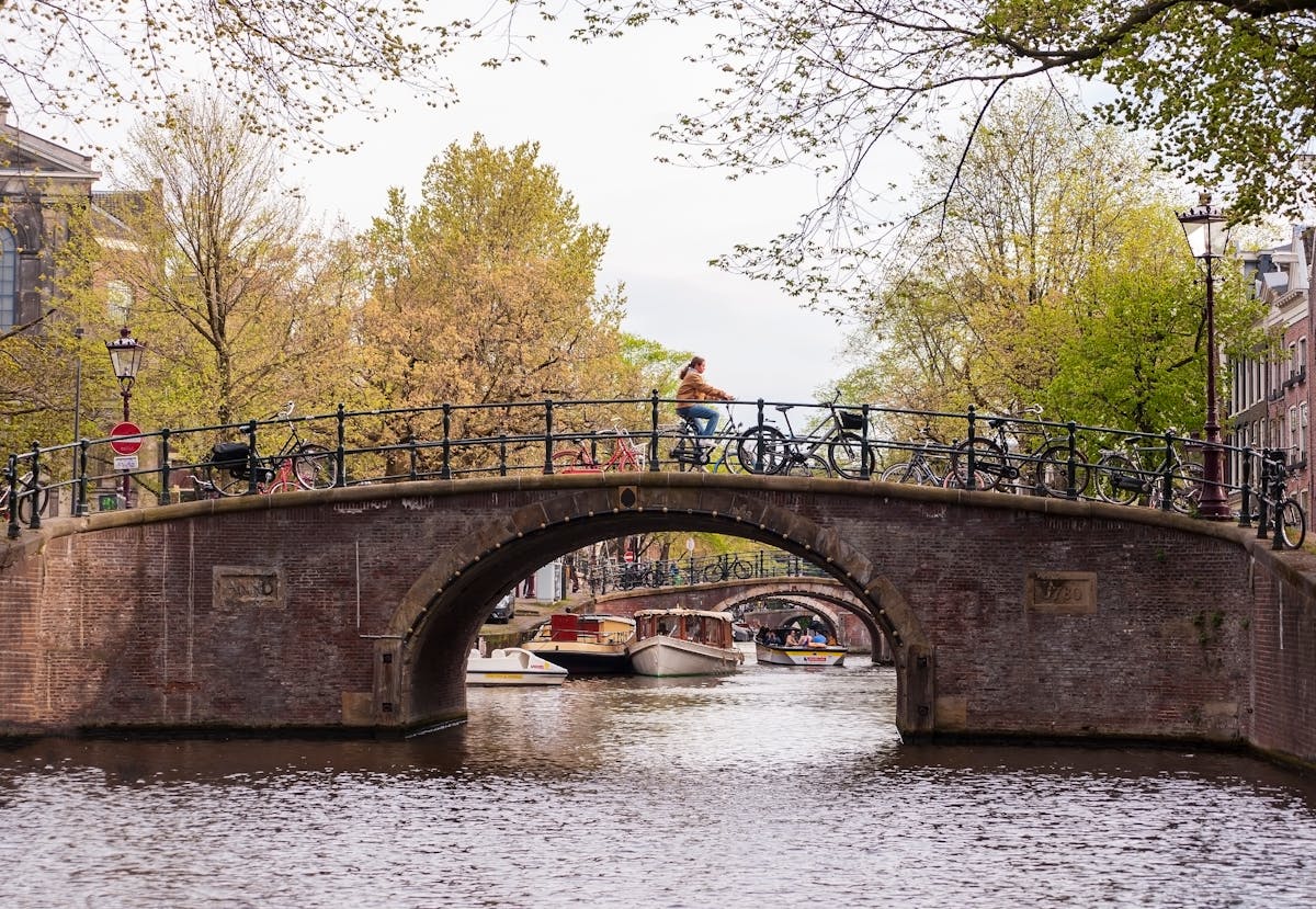Person cycling over a scenic canal bridge in Amsterdam with boats and spring trees