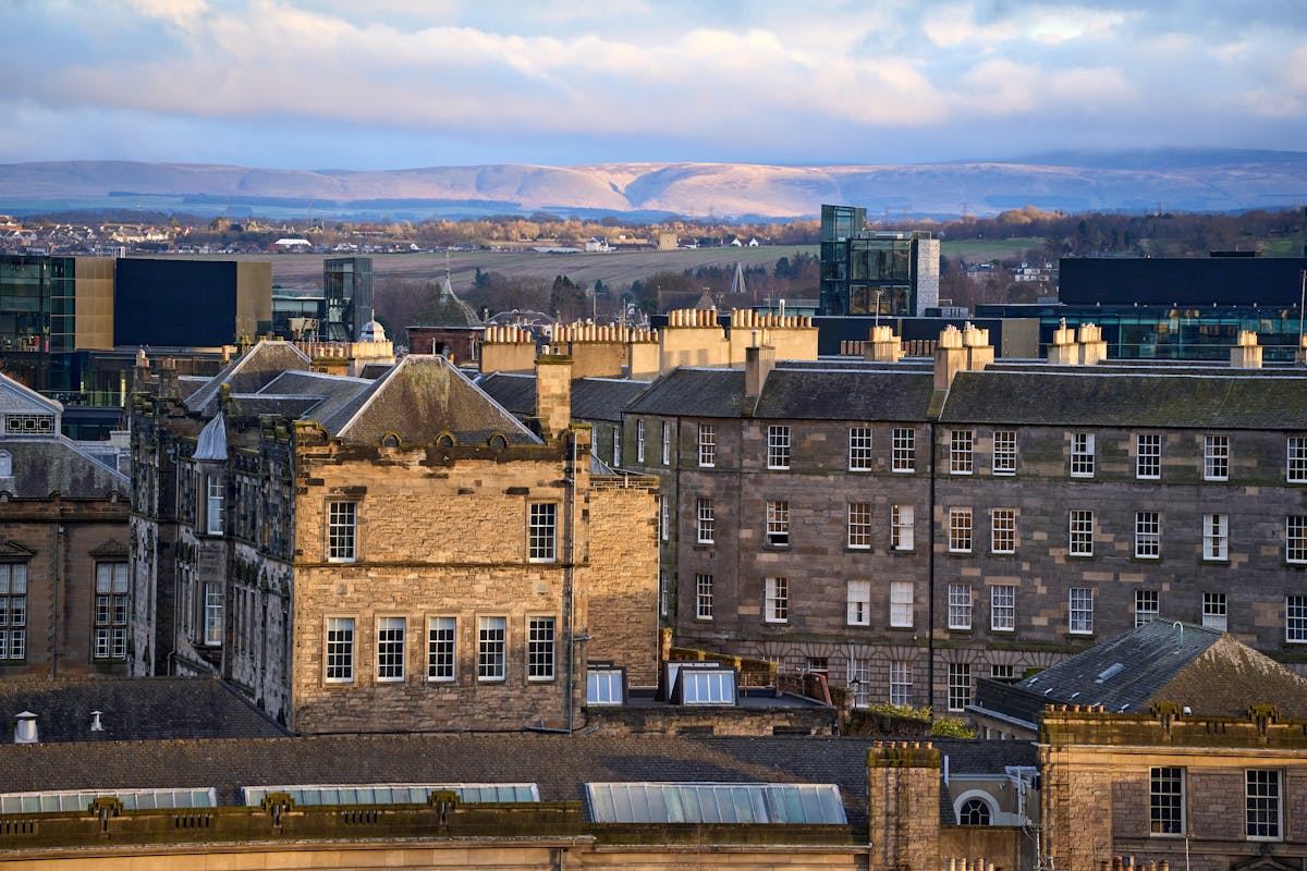 Wide view of Edinburgh historic rooftops and chimneys with distant green hills under a bright sky
