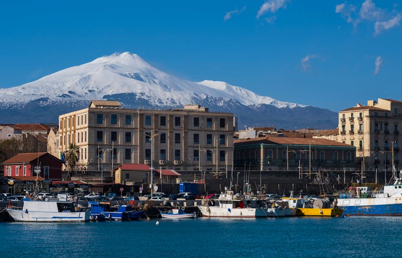 View of Mount Etna with snowy peaks looming over the colorful harbor of Catania Sicily