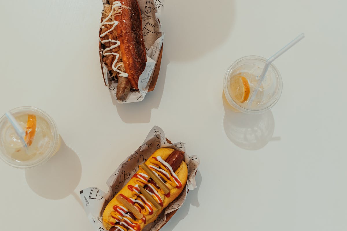 Two gourmet hot dogs with condiments and drinks on a sunny day