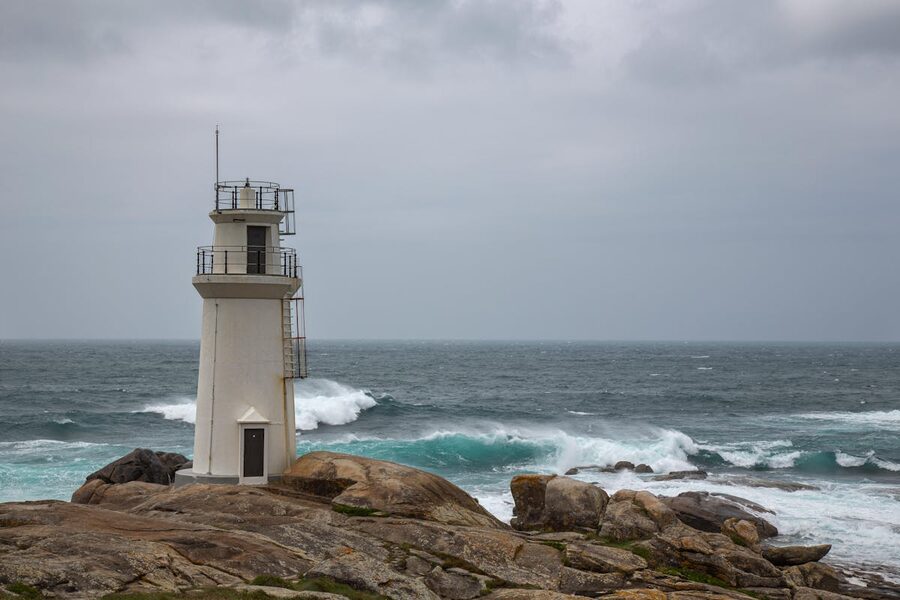 White lighthouse perched on rocky Galician coastline with crashing ocean waves