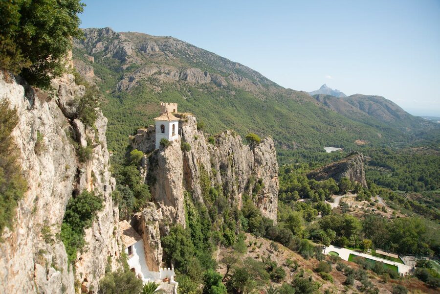 The iconic Guadalest bell tower standing atop a cliff with lush mountains and valley stretching behind it