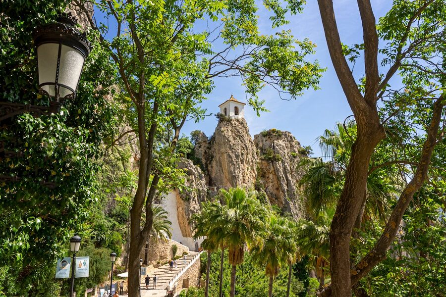 The iconic Guadalest bell tower perched on a rocky cliff surrounded by dense green vegetation and steep mountain walls