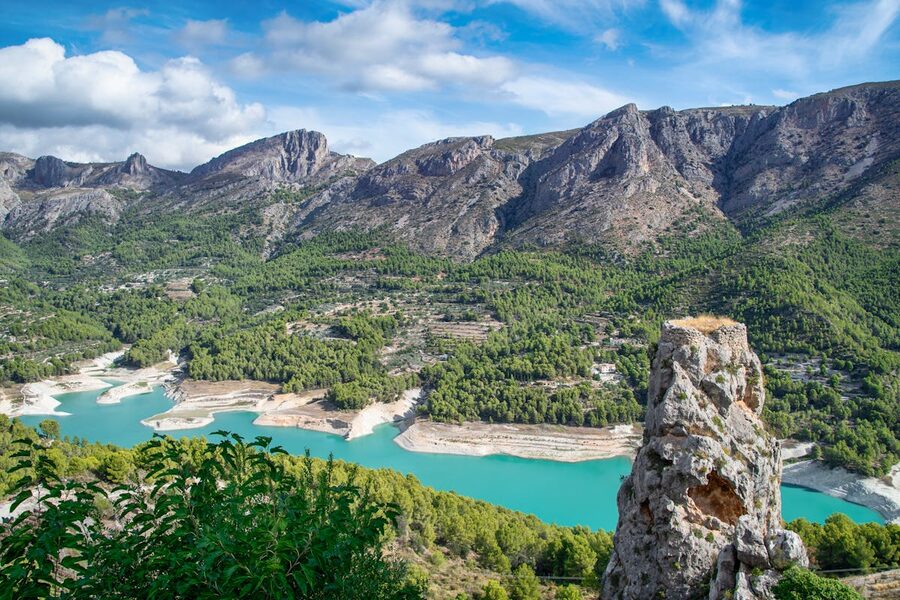 Panoramic view of Guadalest Valley showing turquoise reservoir surrounded by rugged mountains under blue sky