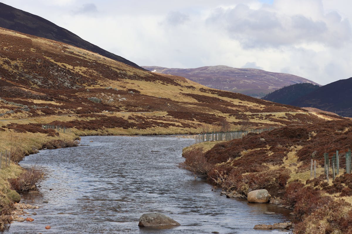 A winding river flowing through the Scottish Highlands with green and brown hills on either side under a grey sky