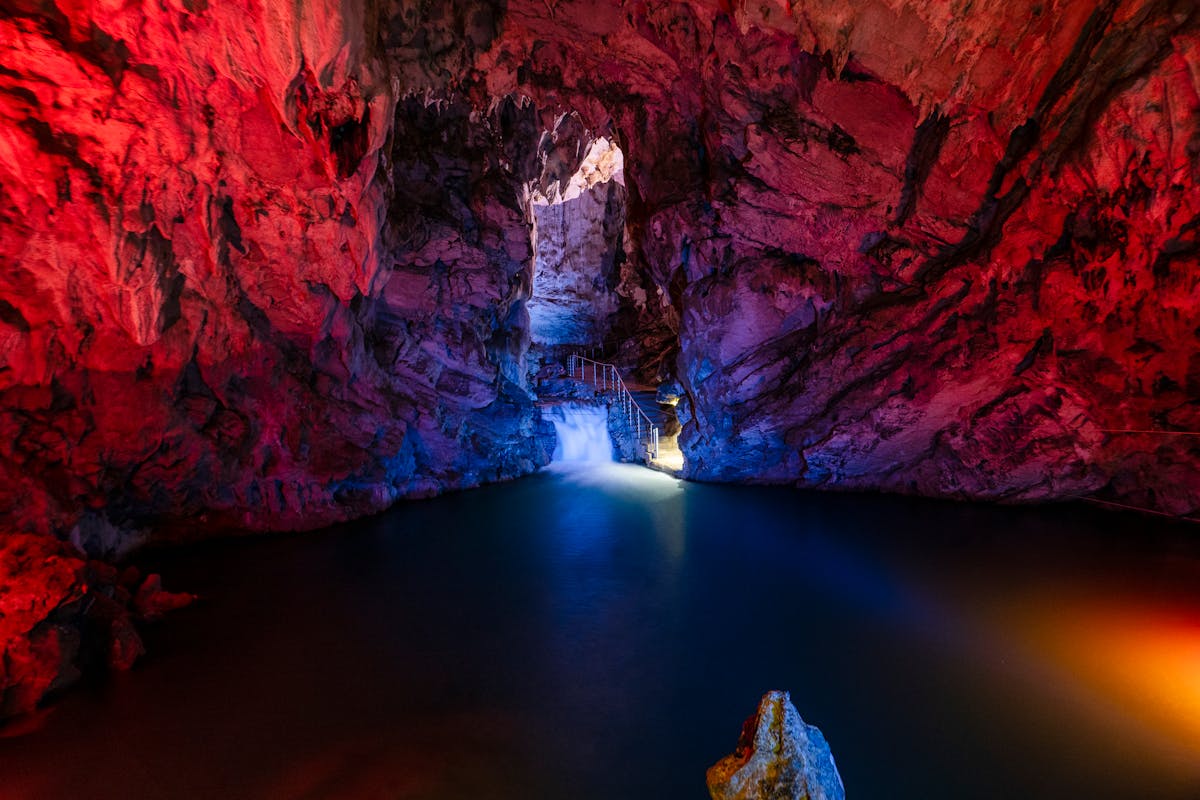 Cave interior illuminated with colorful lighting showing rock formations and a small waterfall