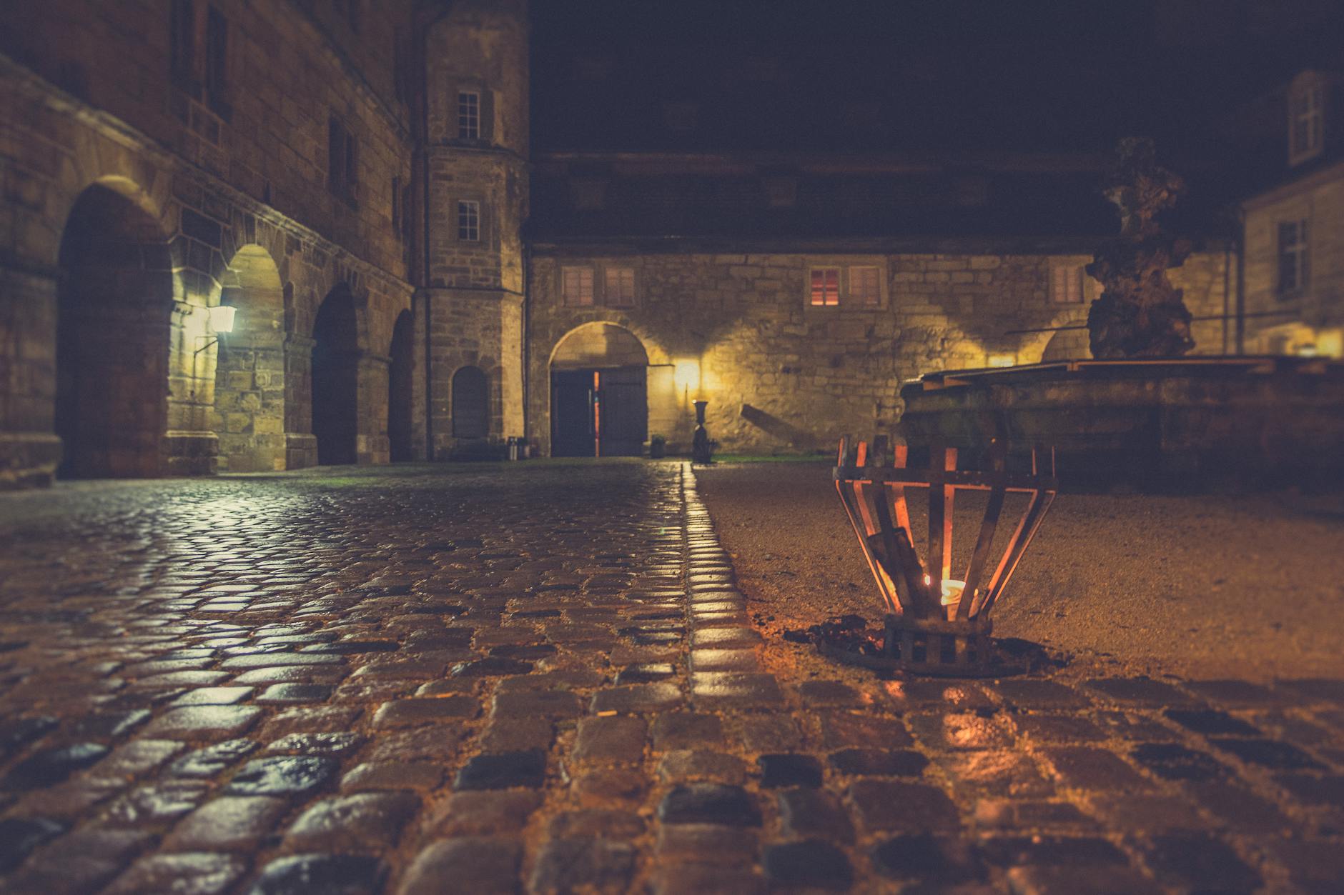 A dimly lit medieval courtyard with cobblestones and a glowing brazier at night