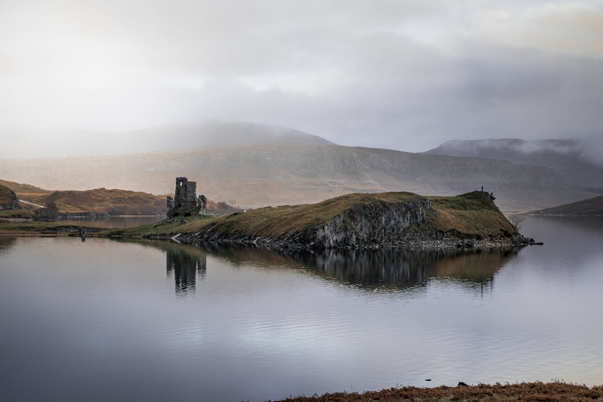 The ruins of Ardvreck Castle reflected on the calm surface of Loch Assynt in Scotland with misty mountains behind