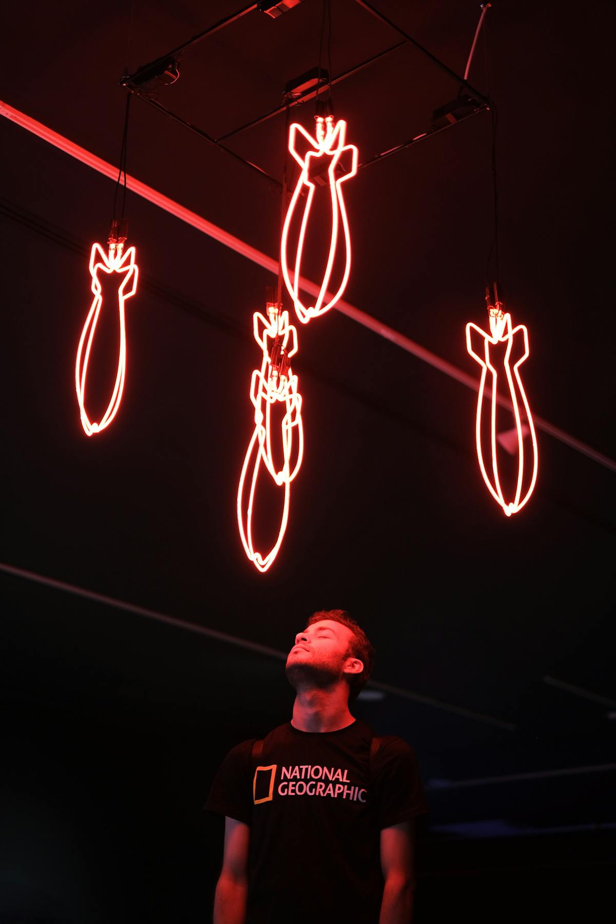 Man standing under red neon bomb lights in a dark room looking upwards pensively