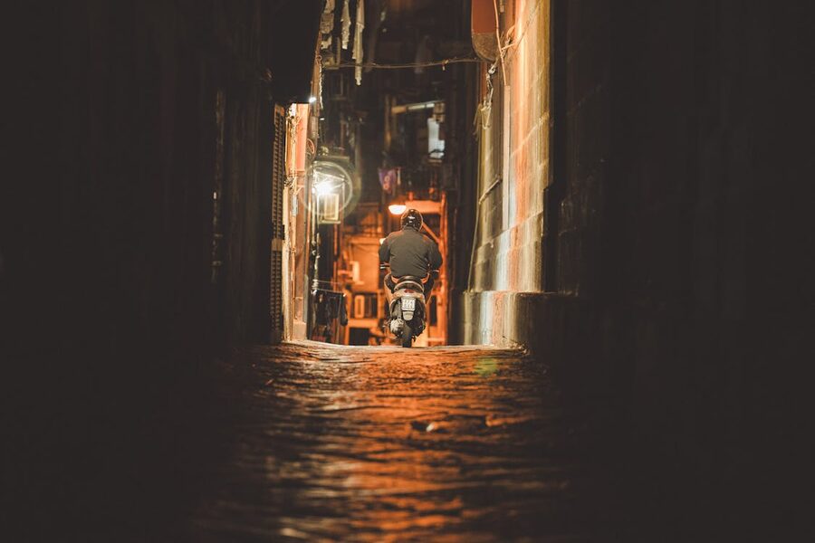 Man riding a motor scooter through a narrow dimly lit alley in Naples at night
