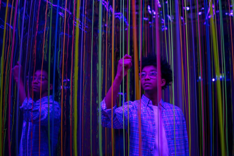 Young person interacts with colorful neon strings in a reflective art installation
