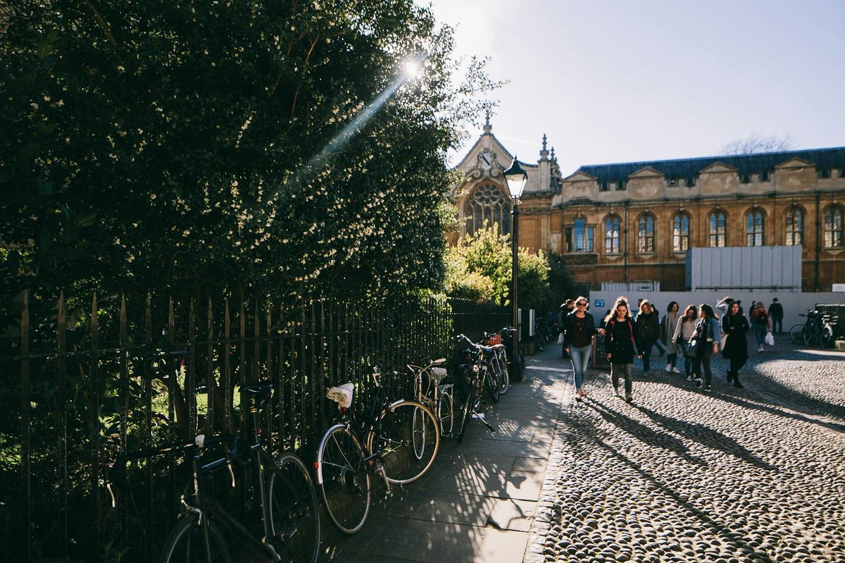 People walking past a sunlit Oxford University building with bicycles