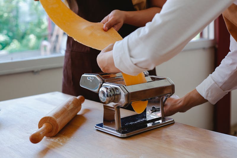 Hands rolling dough through a pasta maker with freshly cut sheets