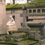 Stone statues and elaborate tombs line a cobblestone path at Pere Lachaise Cemetery in Paris with a crow perched nearby
