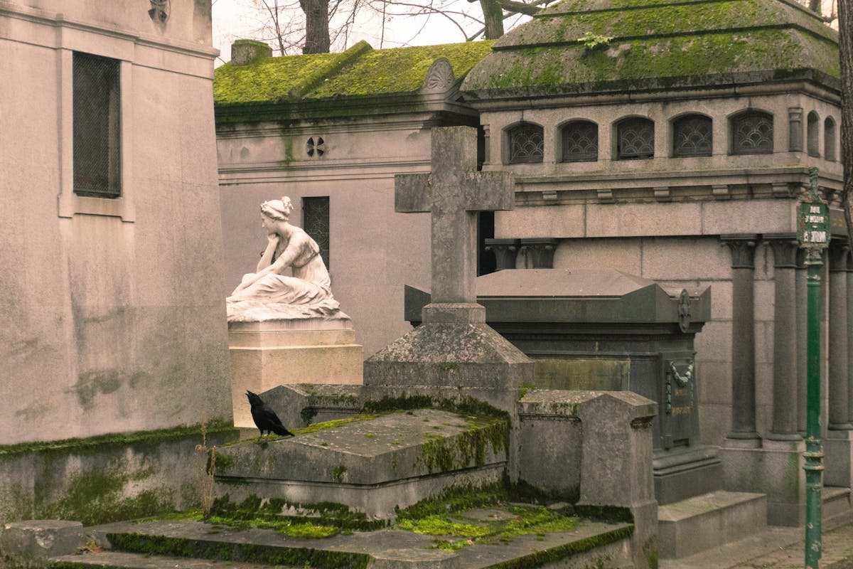 Stone statues and elaborate tombs line a cobblestone path at Pere Lachaise Cemetery in Paris with a crow perched nearby