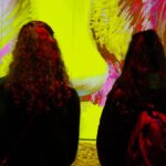 Two visitors with backpacks viewing colorful digital art projections on large walls in an immersive exhibition space