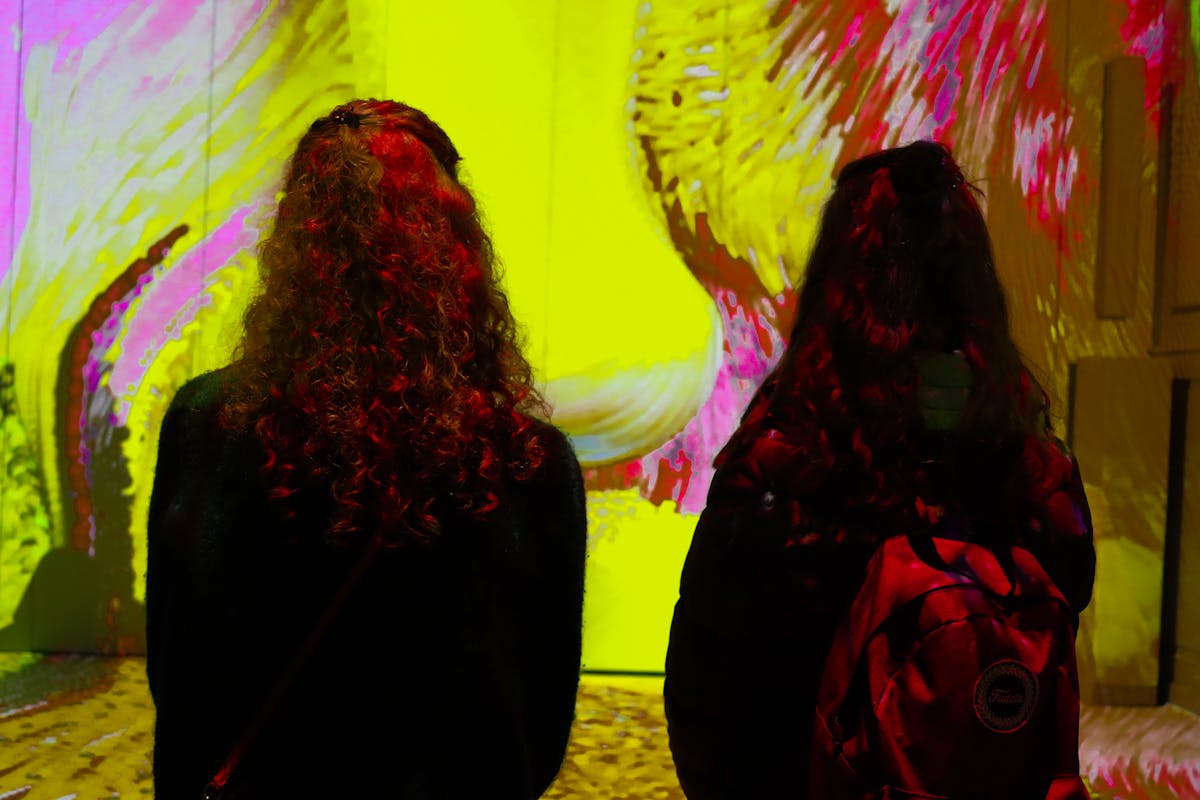 Two visitors with backpacks viewing colorful digital art projections on large walls in an immersive exhibition space