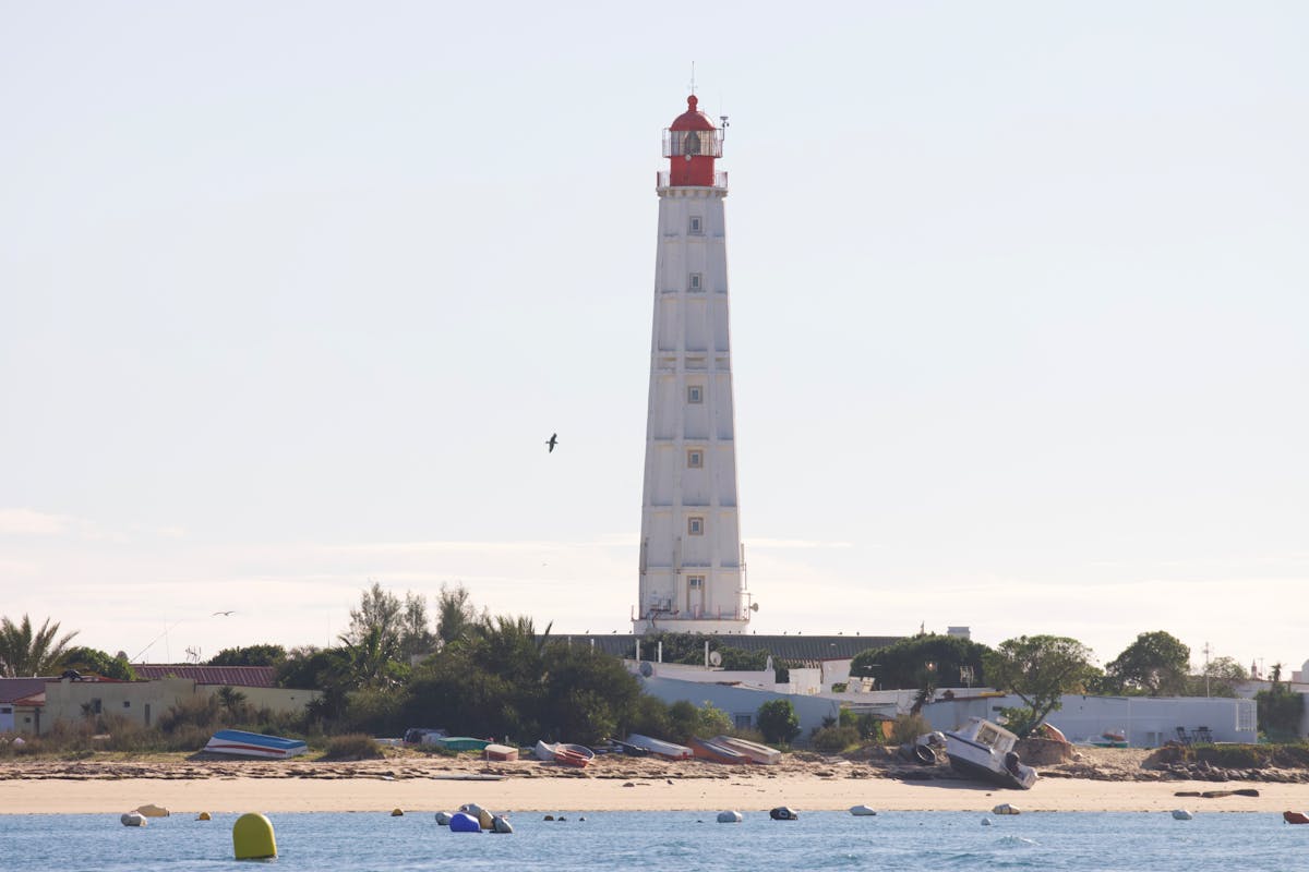 White lighthouse by the sea with colorful boats on the shore in Faro, Portugal