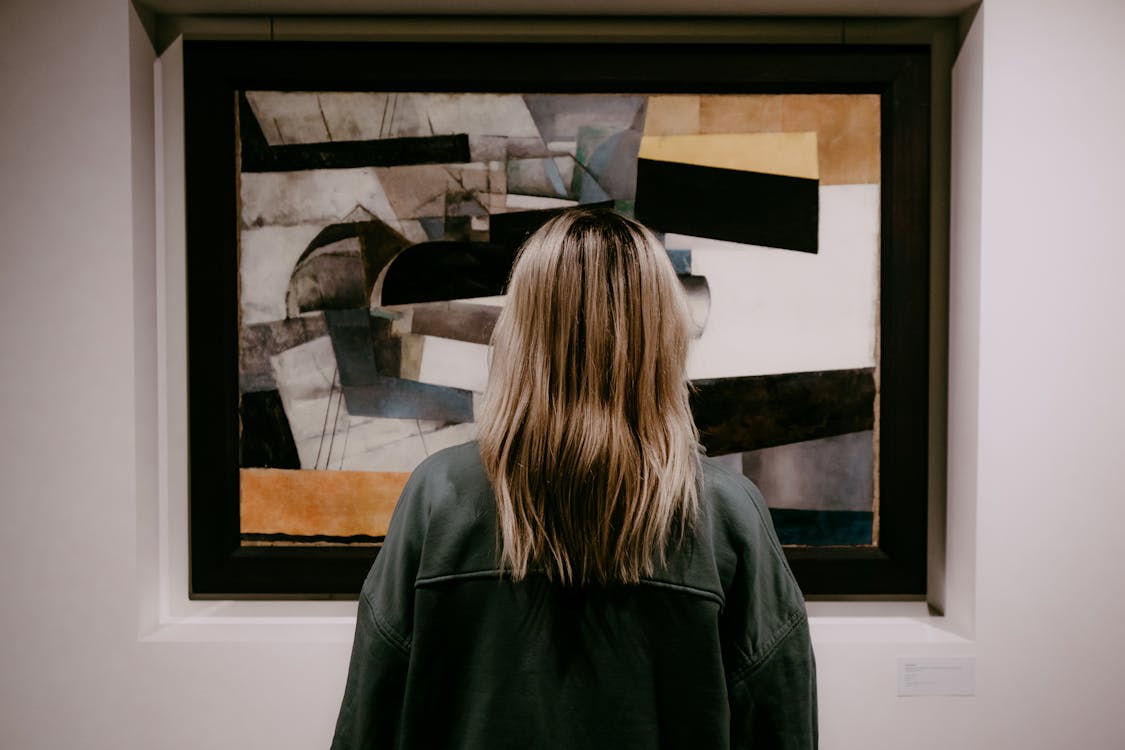 Back view of a woman with blonde hair viewing an abstract painting in an art gallery
