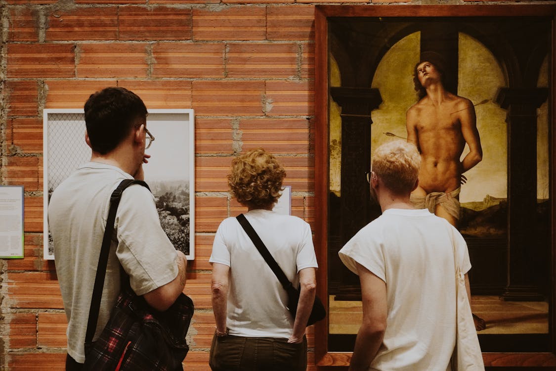 Three people viewing paintings in an art gallery with brick wall backdrop