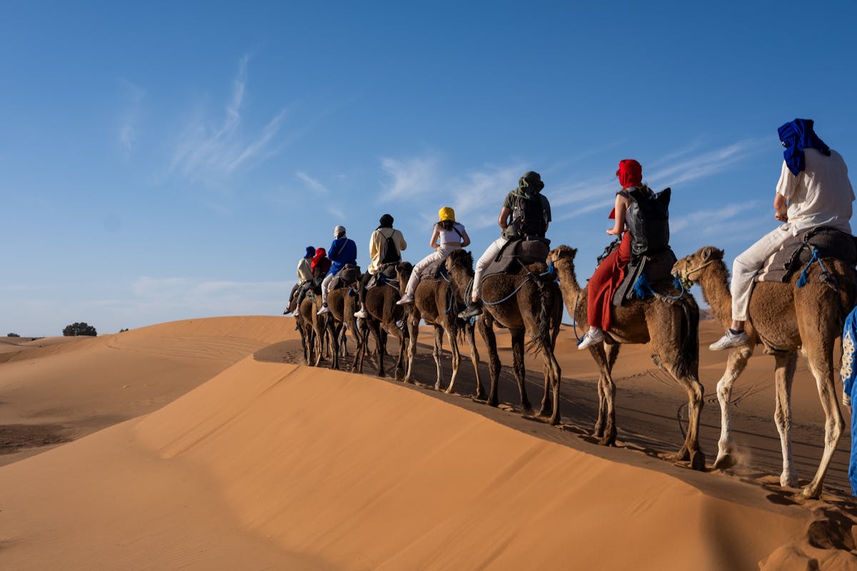 A group rides camels across sandy desert dunes under clear blue sky