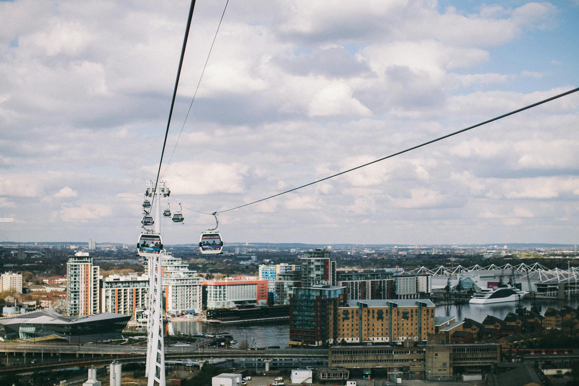 Multiple cable car gondolas suspended on cables above the River Thames with London buildings and cloudy sky