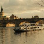 Cruise boat sailing under Charles Bridge on the Vltava River in Prague during golden hour