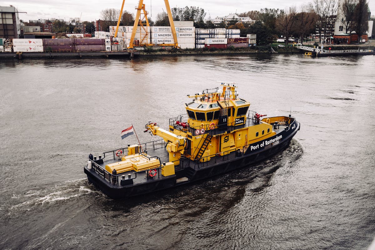 A yellow tugboat sails at the Port of Rotterdam with container ships and cranes in the background