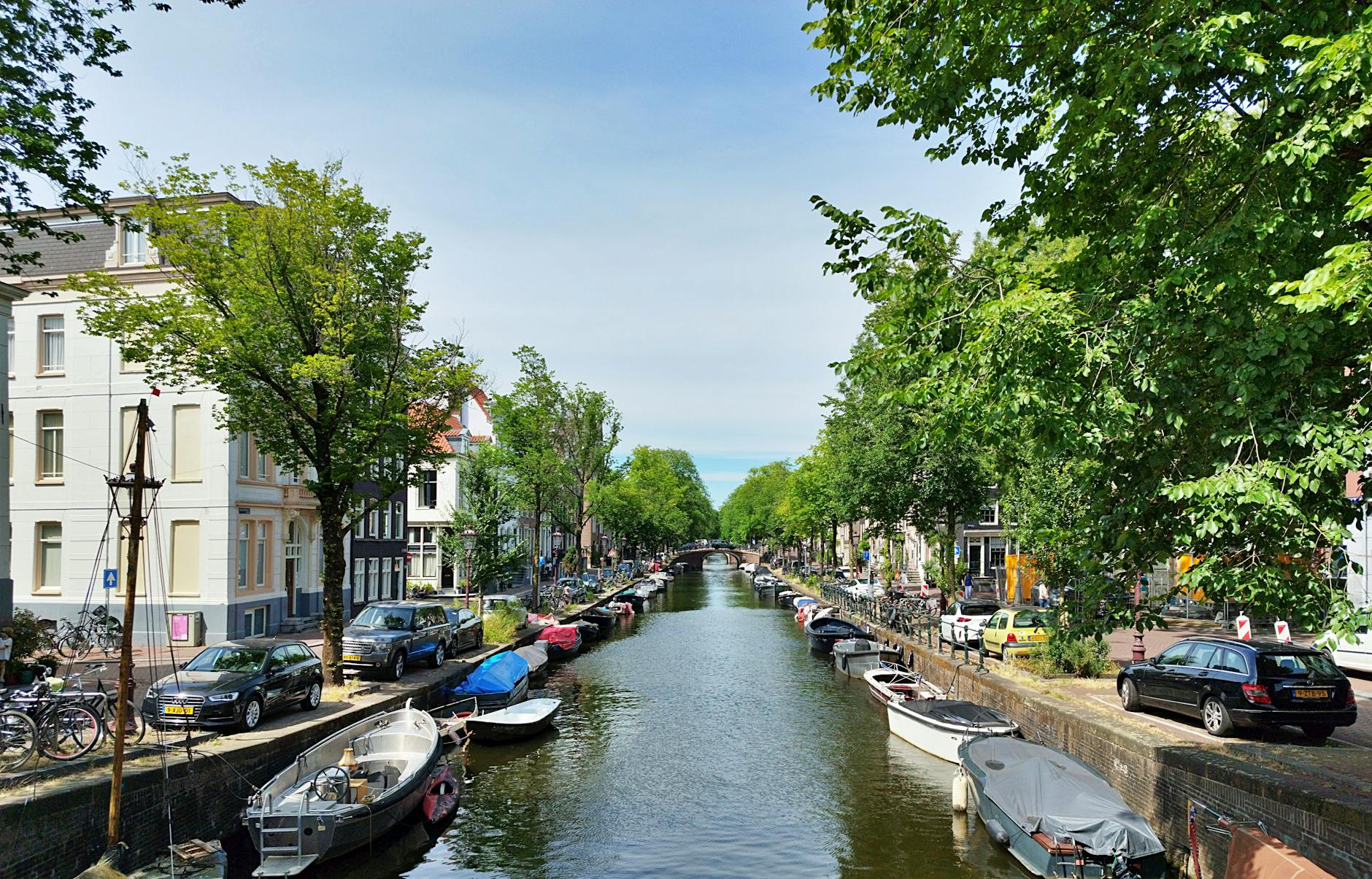 A picturesque Amsterdam canal lined with boats cars and lush green trees under a clear sky