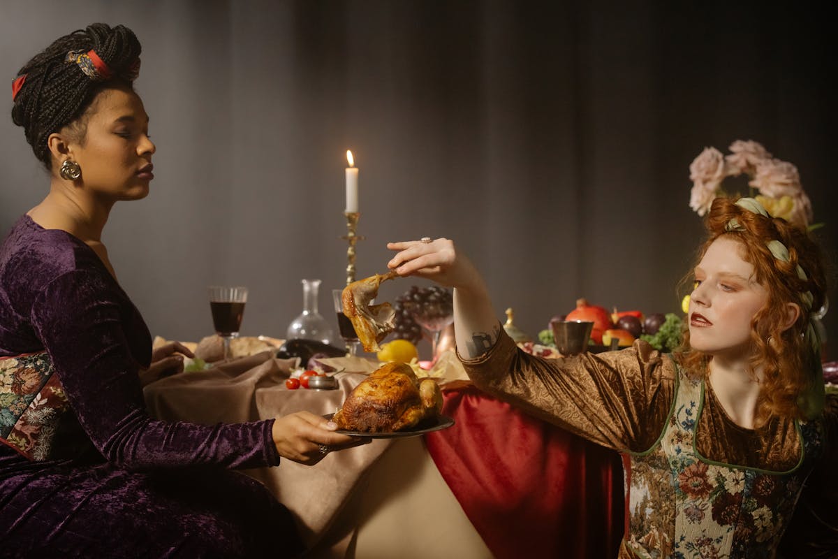 Two women in renaissance costumes at a candlelit feast with food and flowers