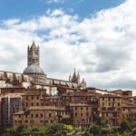 Siena Cathedral and bell tower rising above the historic terracotta rooftops of Siena under a cloudy sky