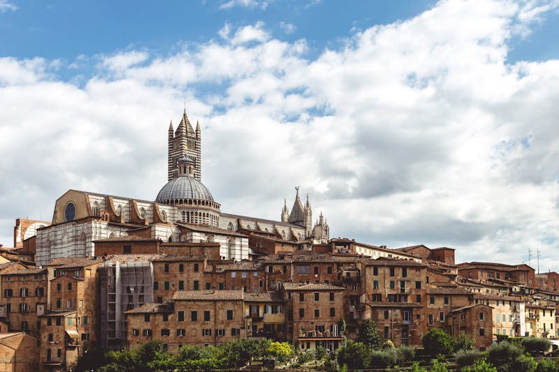 Siena Cathedral and bell tower rising above the historic terracotta rooftops of Siena under a cloudy sky