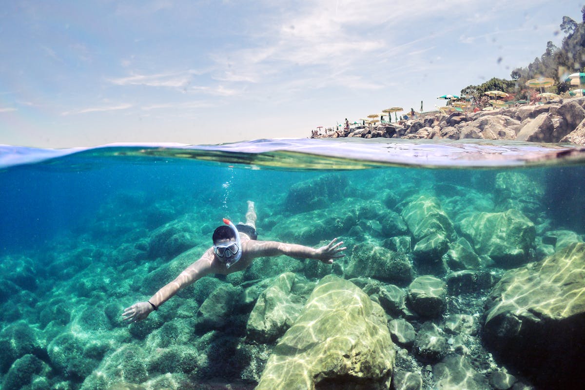A snorkeler swimming underwater near a rocky coastline with clear water