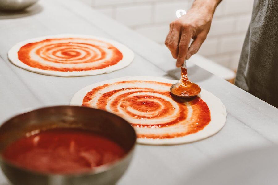 Hand spreading tomato sauce on pizza dough in an indoor setting
