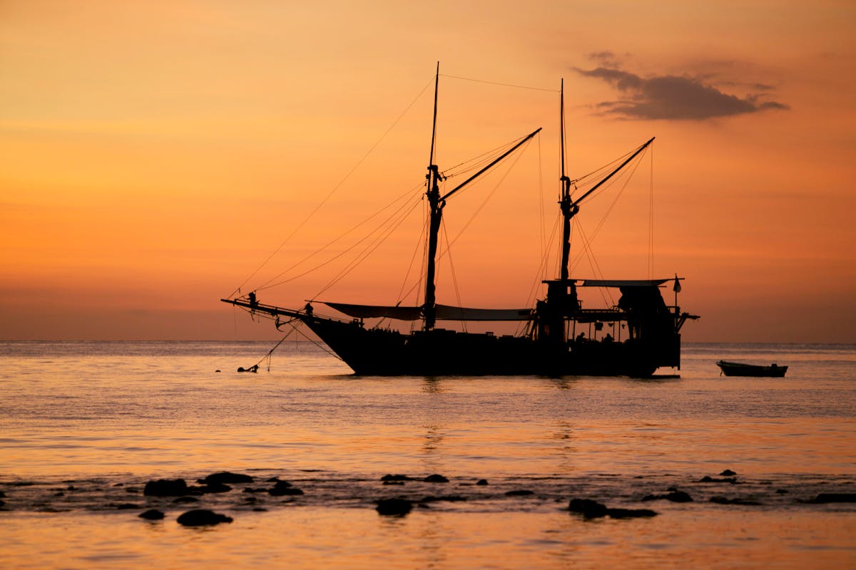 Silhouette of a classic sailboat against a bright orange sunset over calm water