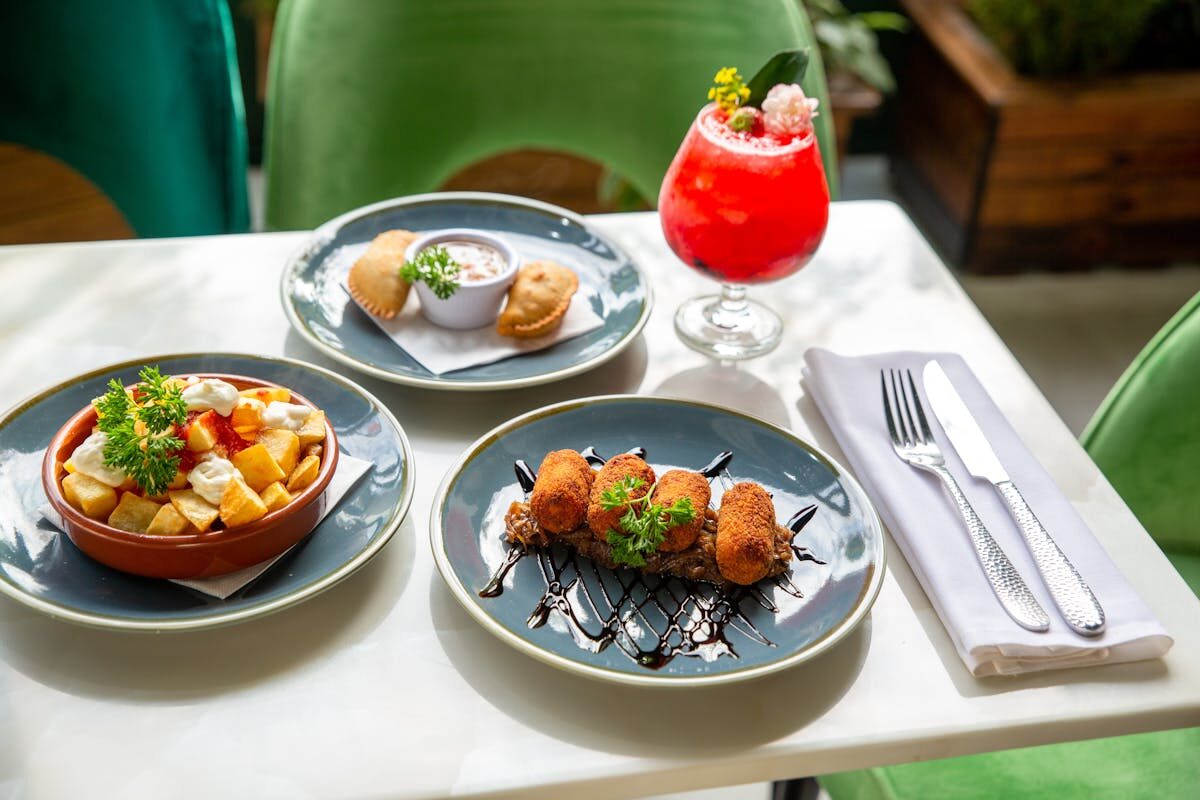 A spread of various Spanish tapas dishes with a cocktail on a sunlit restaurant table
