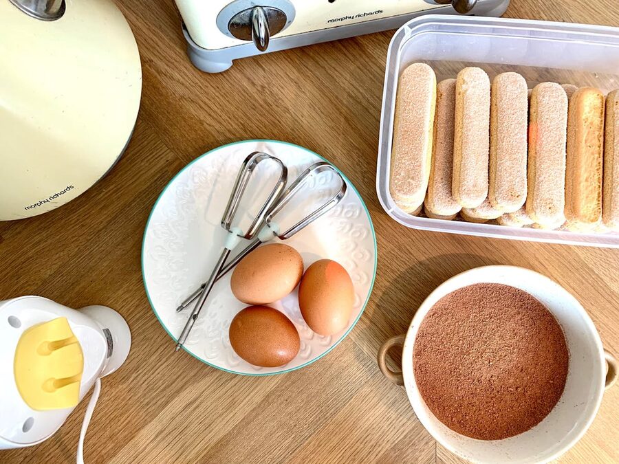 Top-down view of tiramisu ingredients including eggs cocoa powder and ladyfingers on a wooden surface