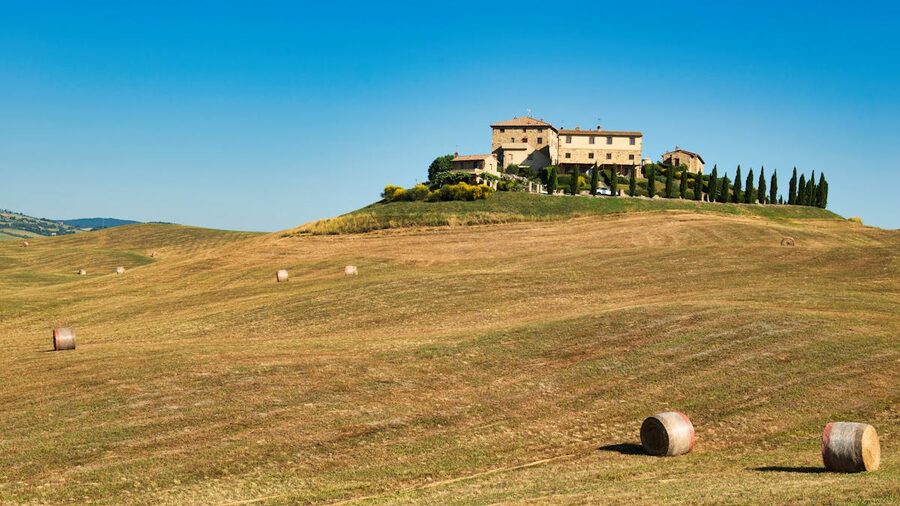 A picturesque Tuscan farmhouse atop rolling hills surrounded by hay bales under dramatic sky