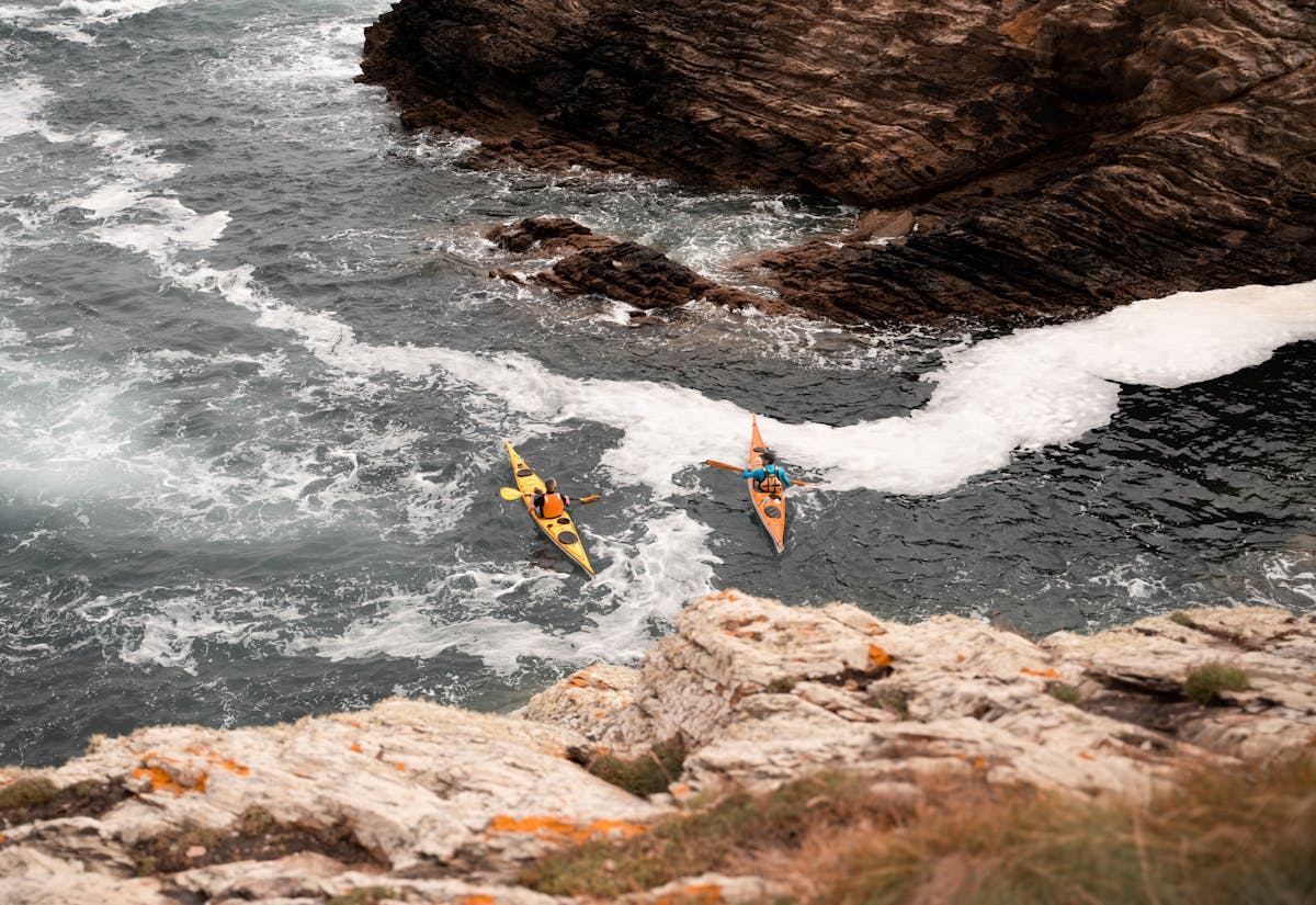 Two kayakers paddling through waves near rocky cliffs